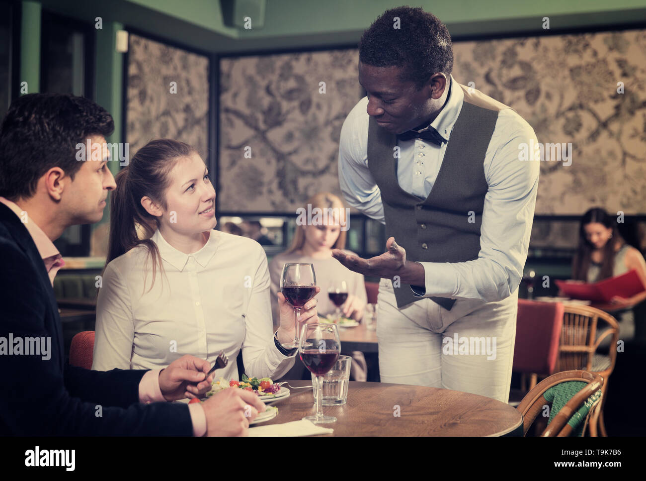 Positive elegant couple talking with polite African American waiter ...