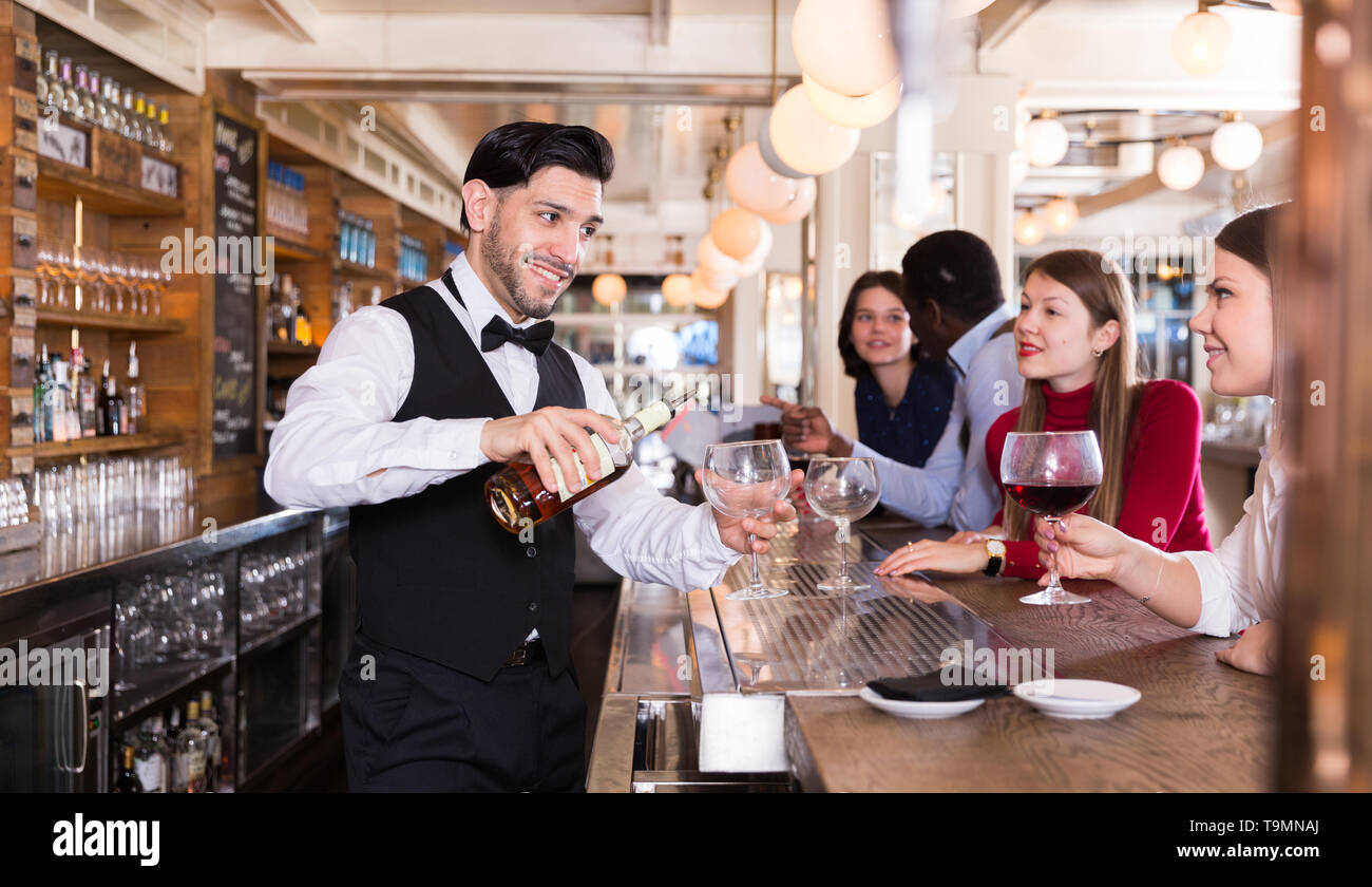 Smiling bartender pouring beverages and talking to cheerful visitors in ...
