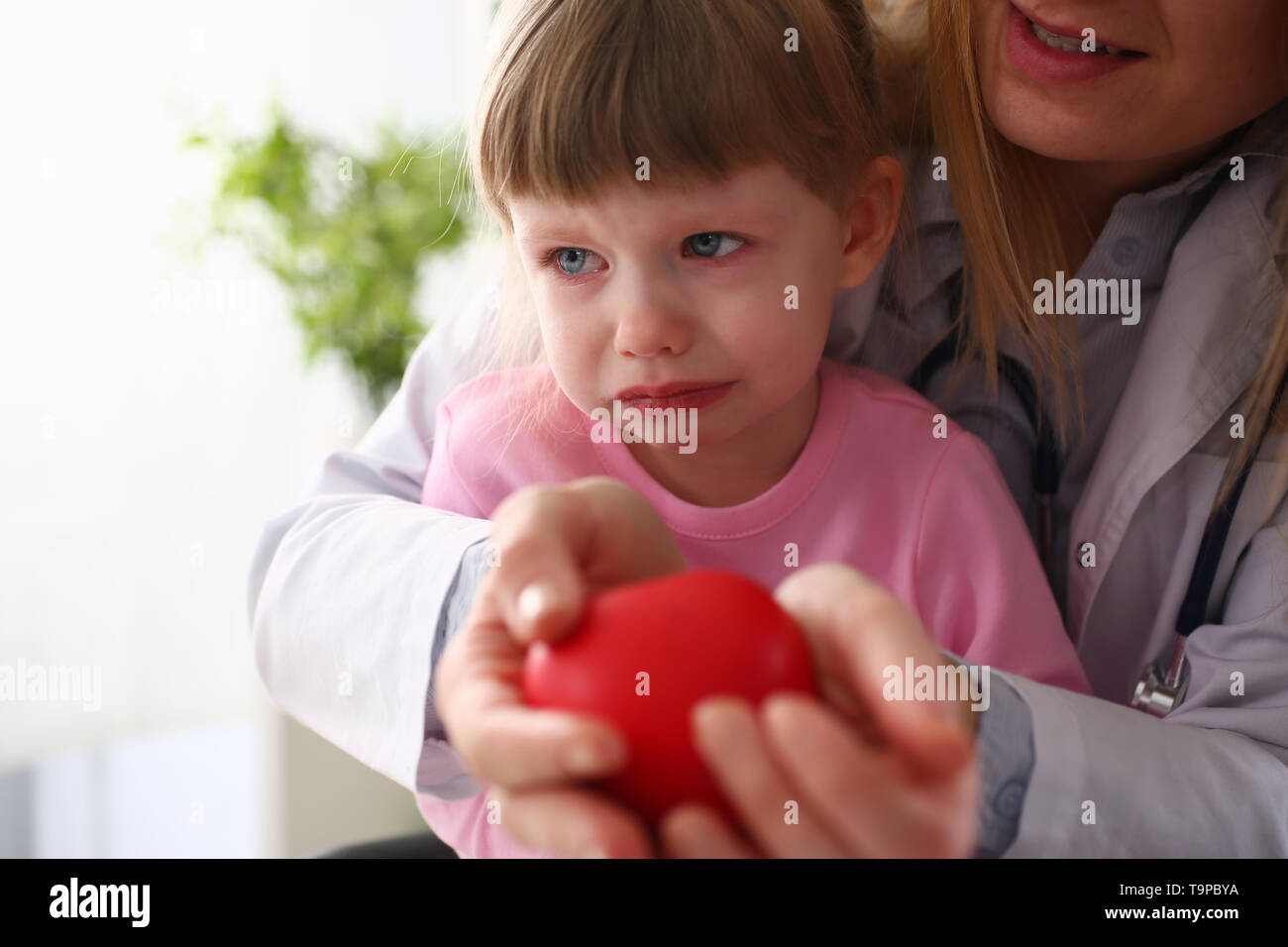 Scared little baby girl visiting doctor holding in hands red toy heart ...