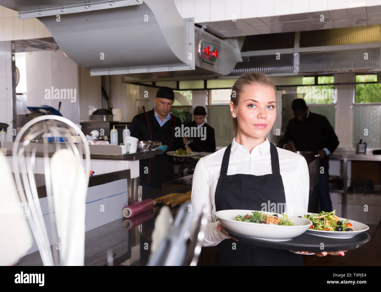 Portrait of attractive waitress with ordered dishes in kitchen of ...