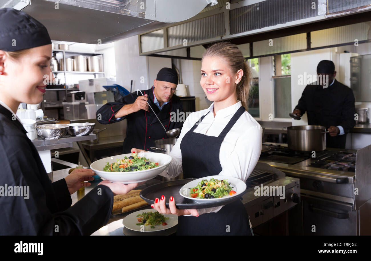 Successful young waitress taking cooked ordered meals from female chef ...