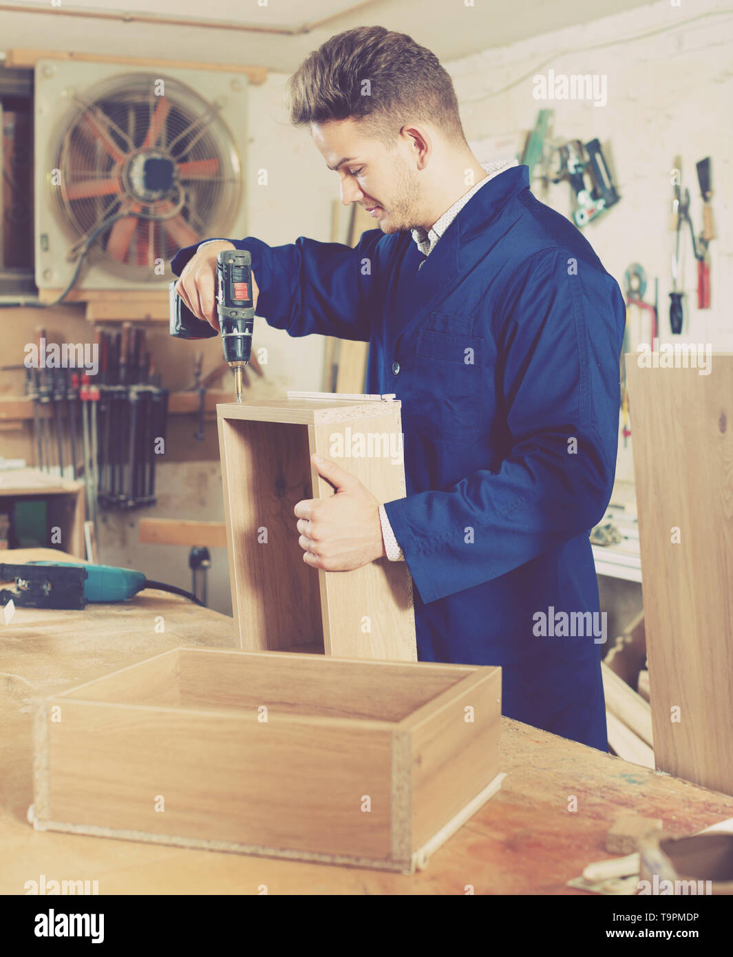 Young smiling guy using drilling machine to make drawer at workshop ...