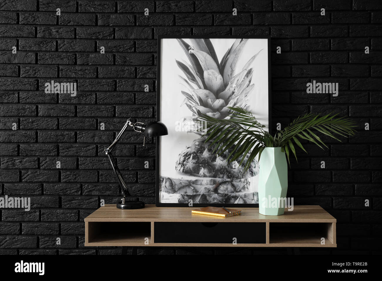 Vase with tropical leaves, picture and lamp on table near dark brick ...