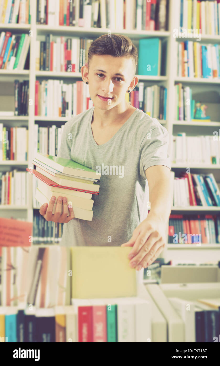 Positive teenager boy looking for new book on shelves in book store ...