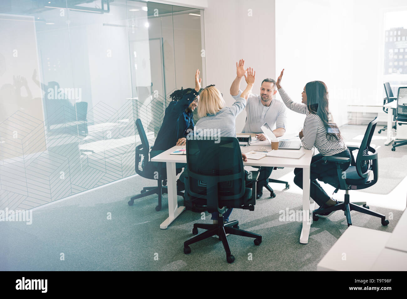 Smiling group of diverse work colleagues sitting around an office table and high fiving each ...