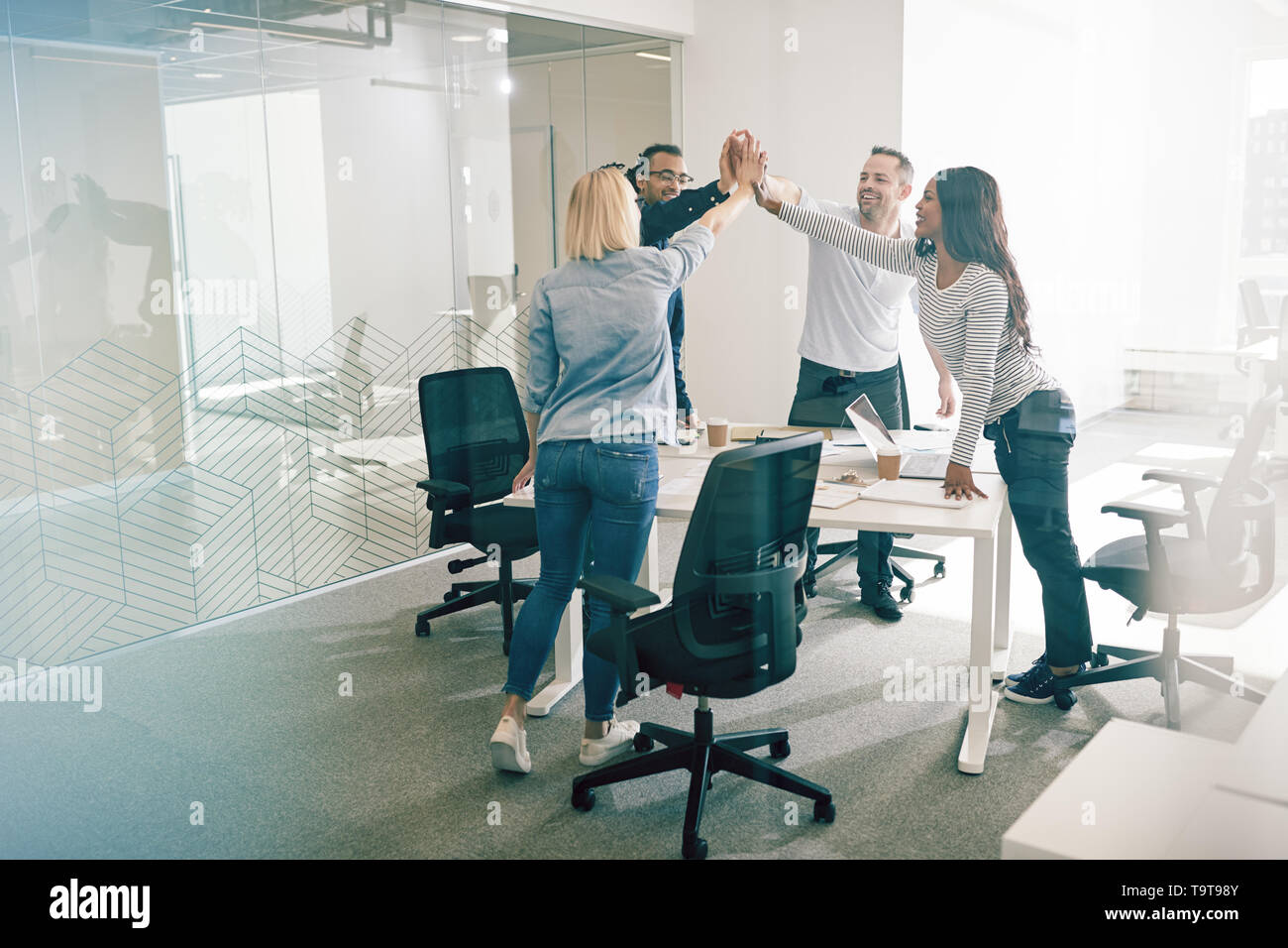 Diverse group of smiling work colleagues standing around an office table and high fiving each ...