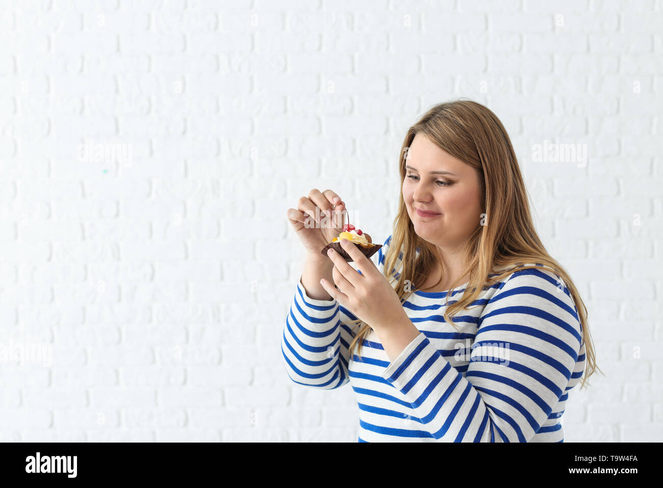 Happy plus size girl with sweet cake on white brick background. Concept ...