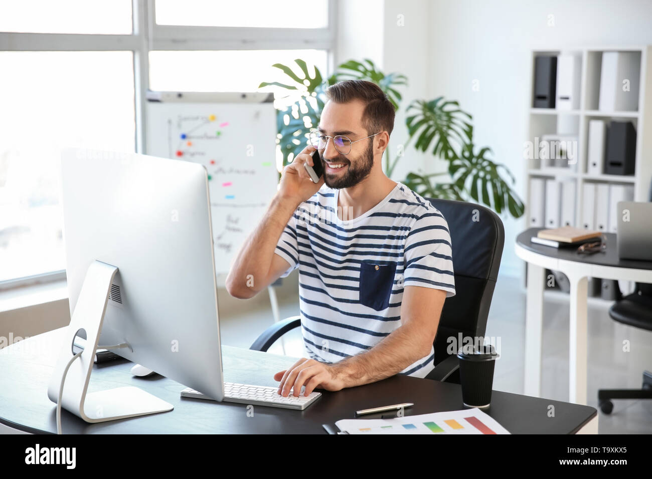 Young man talking on mobile phone while working on computer in office ...