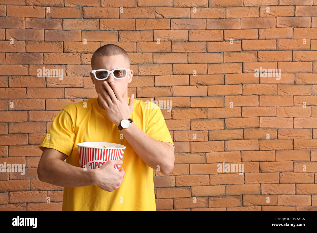 Scared young man with popcorn watching horror movie against brick wall ...