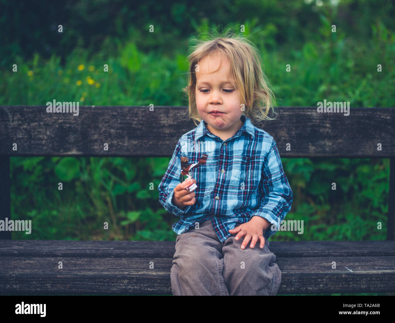 A little toddler is sitting on a bench in nature and is eating a fruit ...