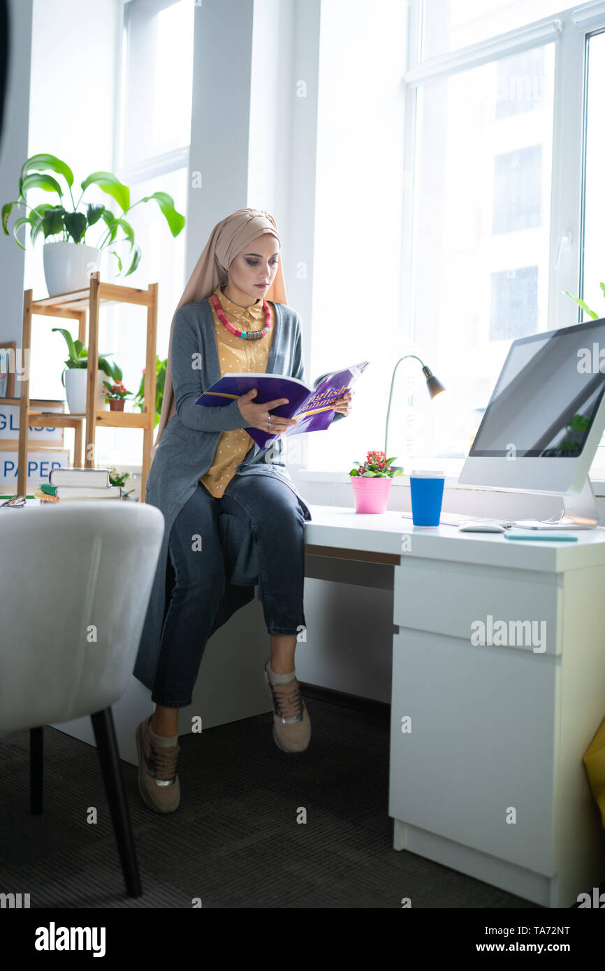 Stylish Muslim teacher sitting near computer and reading book Stock ...