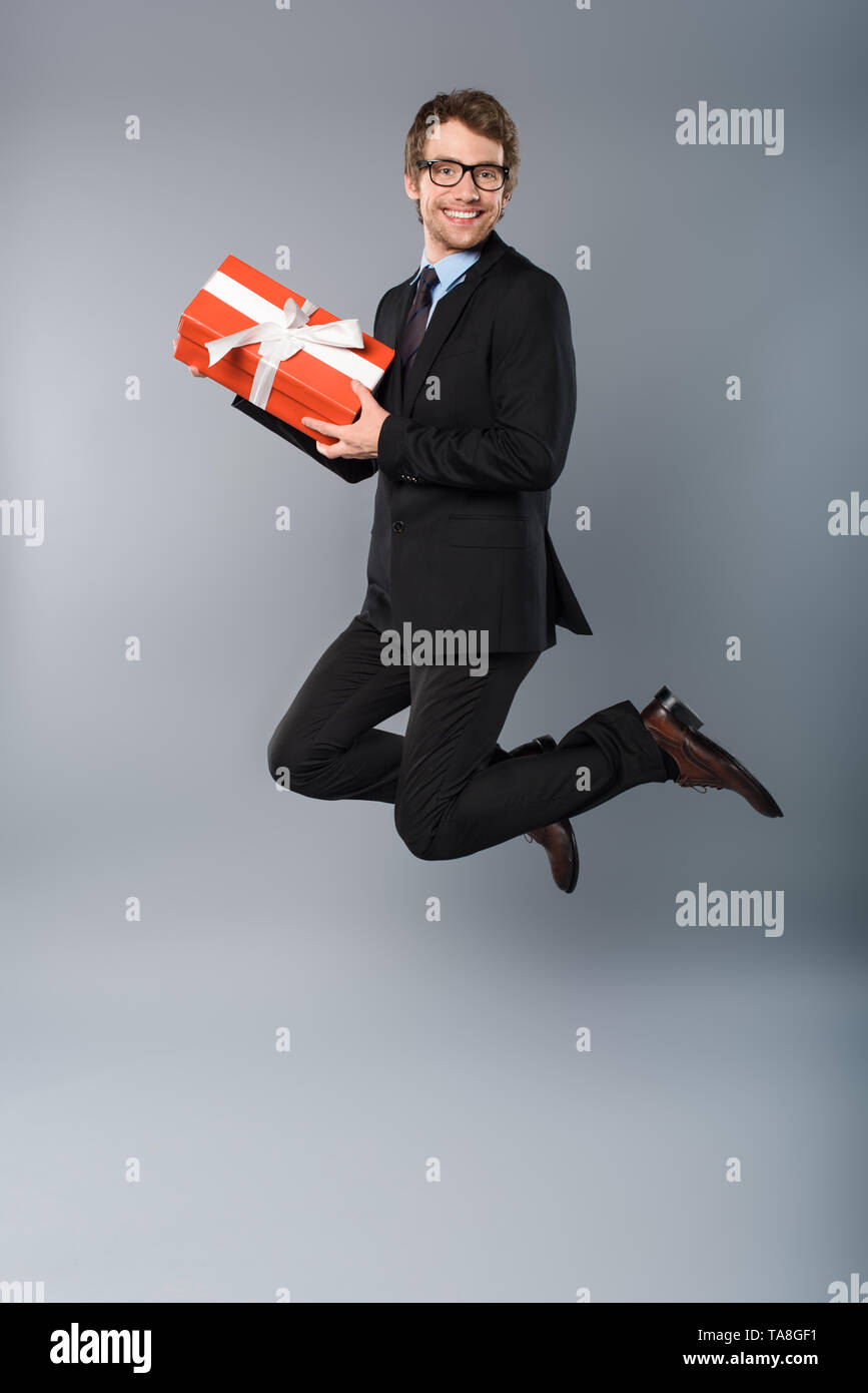 happy businessman holding present while jumping on grey background ...