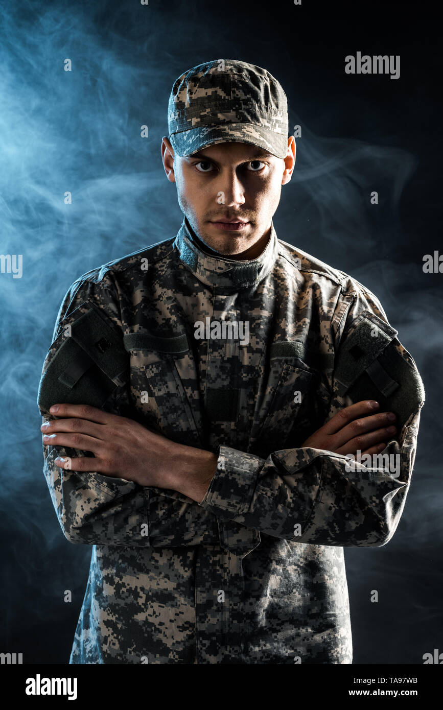 confident soldier in military cap and uniform standing with crossed ...