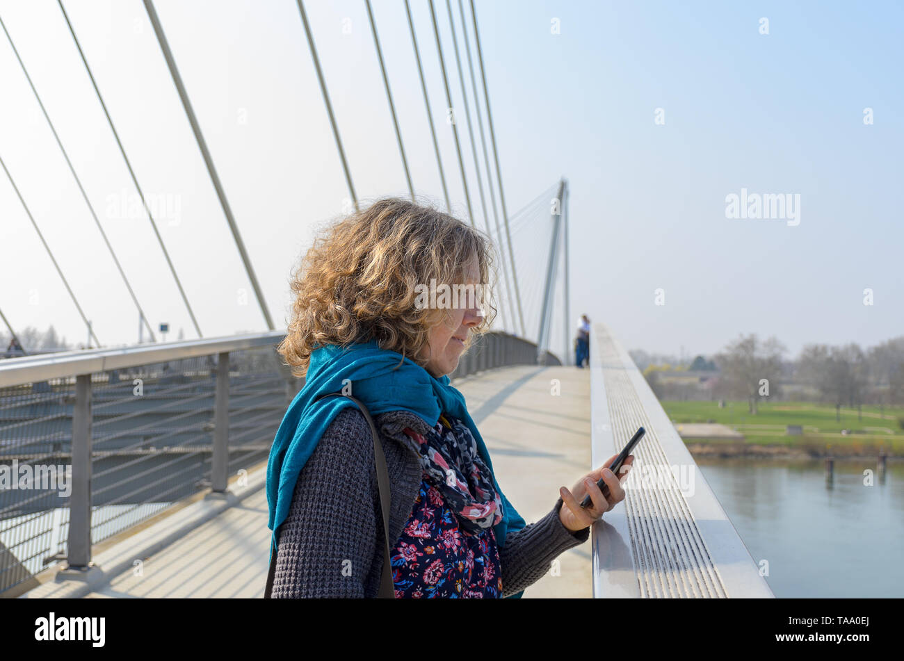 Woman with curly hair standing on modern pedestrian bridge with cell ...