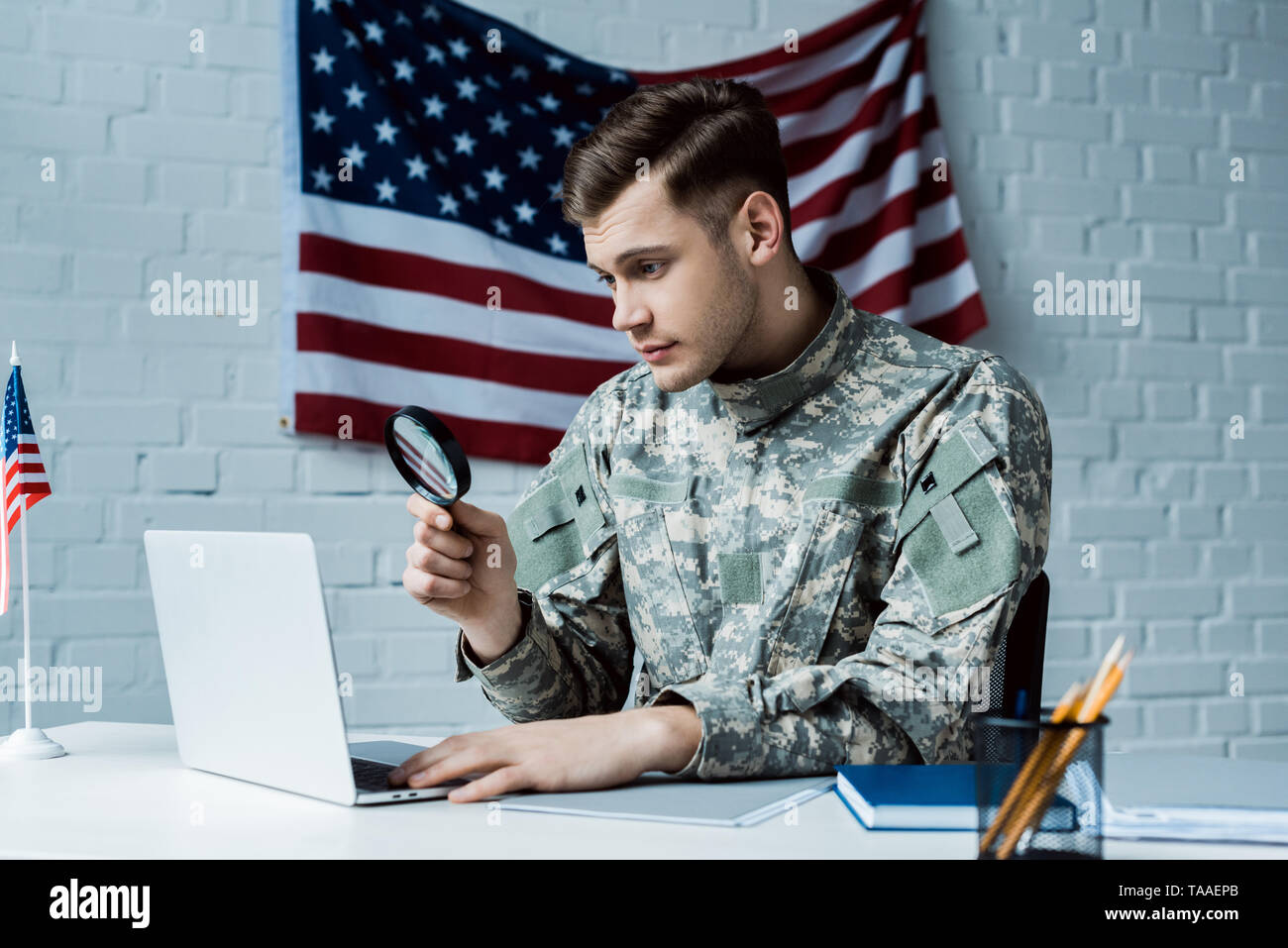 young soldier in uniform holding magnifying glass near laptop in office ...