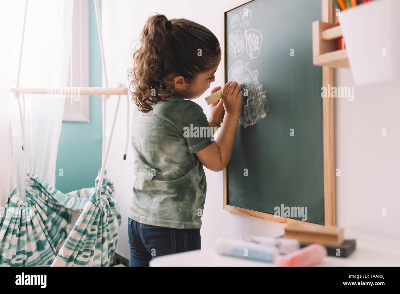 little girl drawing with a chalk on a chalkboard at her room at home ...
