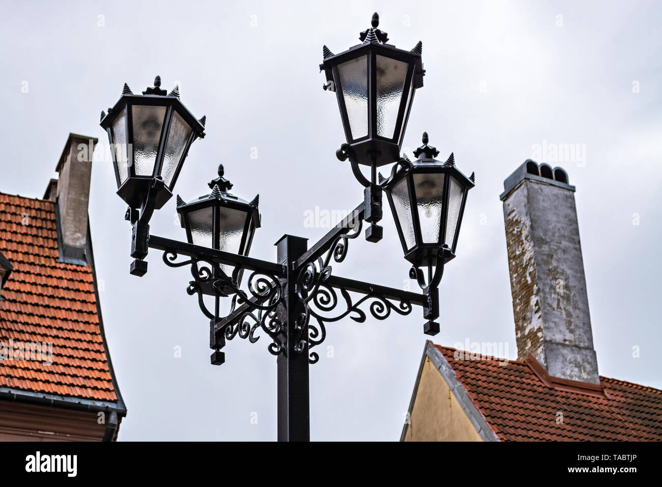 Four street lamps on one pole against a gray sky, between two roofs of ...