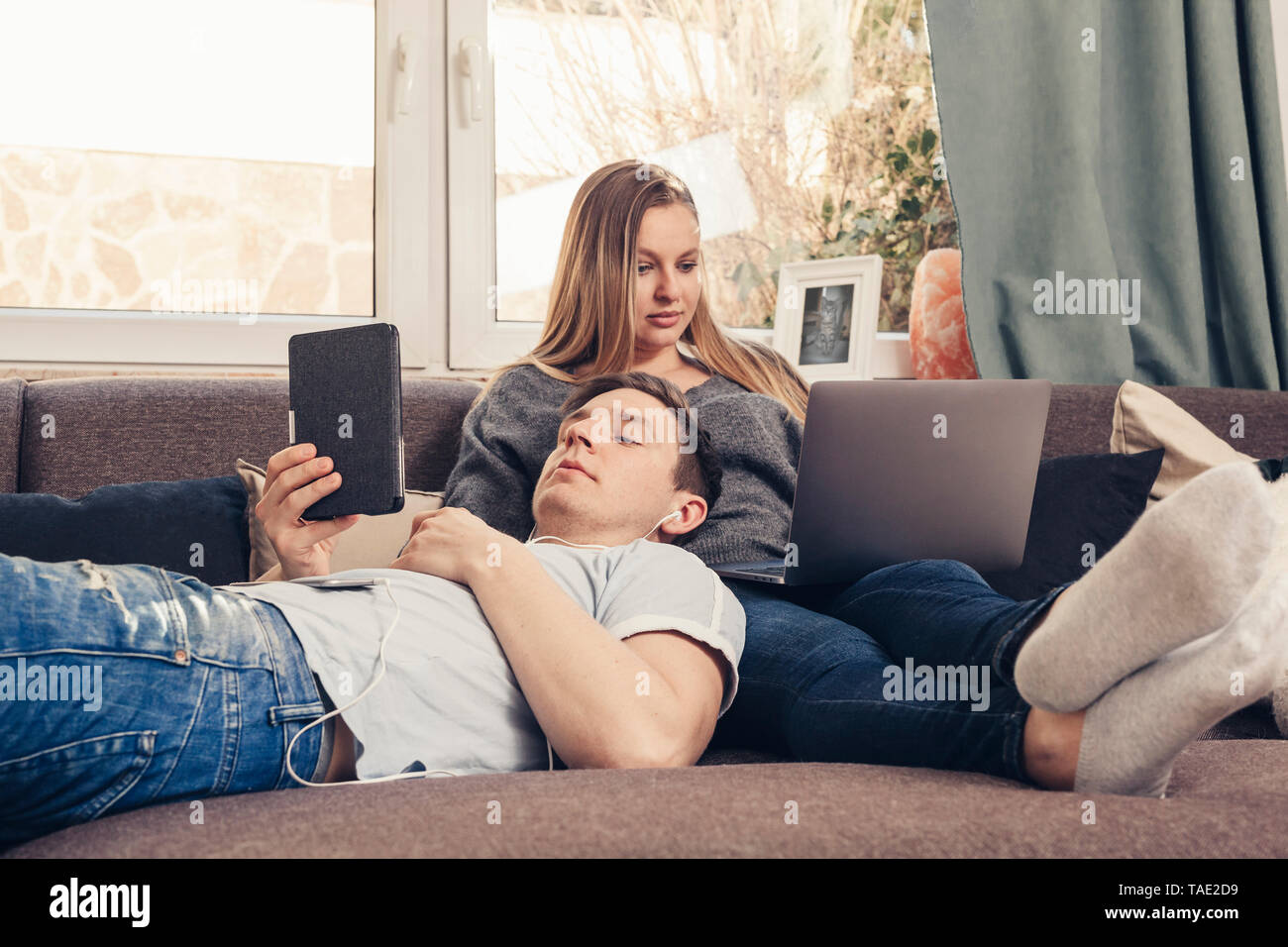 Young couple relaxing on sofa and using notebook, smartphone and ebook ...