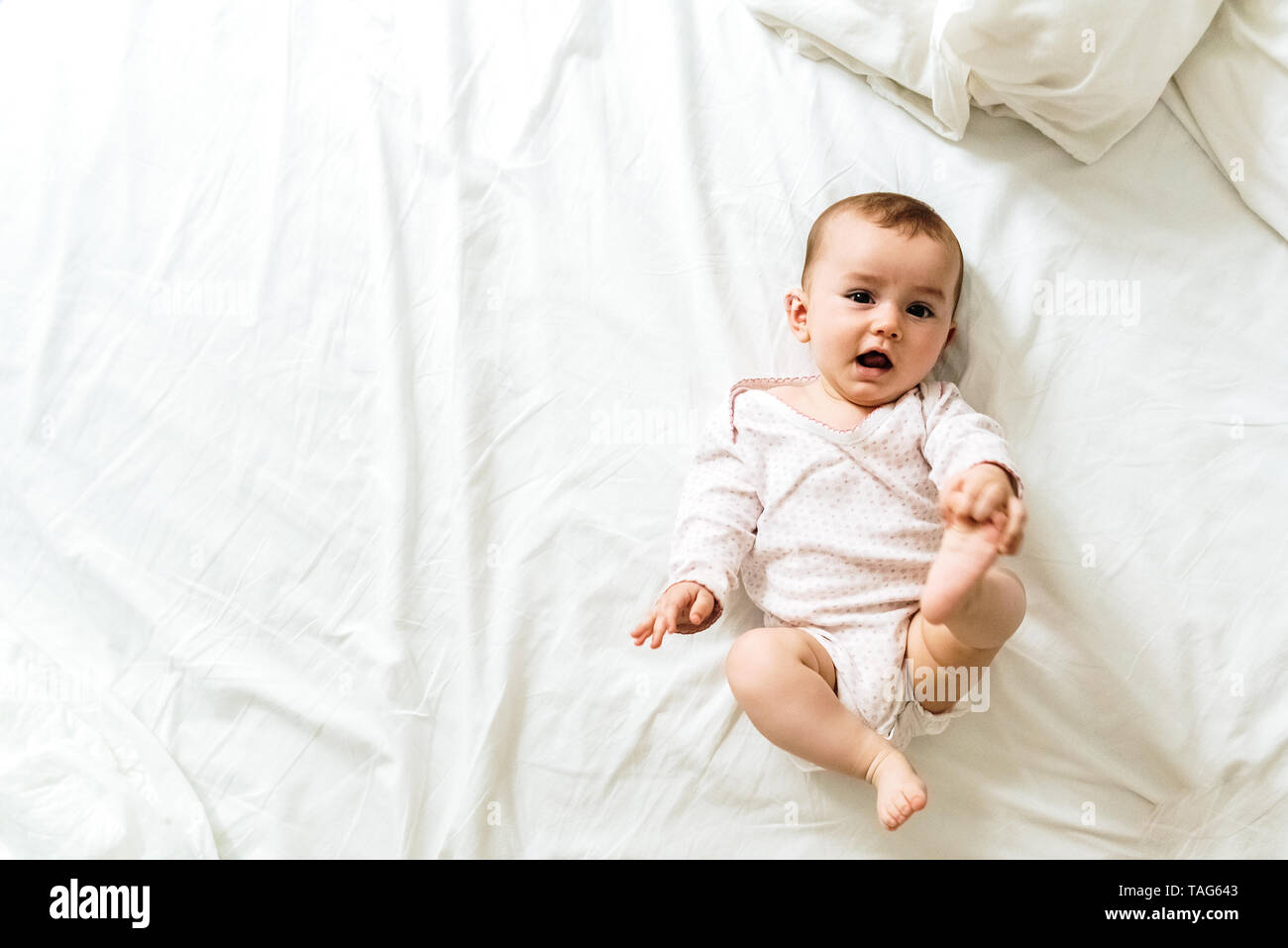 Happy and smiling adorable 6 month old baby girl lying on a bed, lifestyle isolated on natural ...