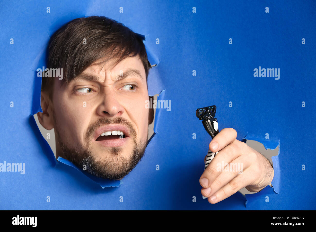 Displeased man with razor visible through holes in torn color paper ...