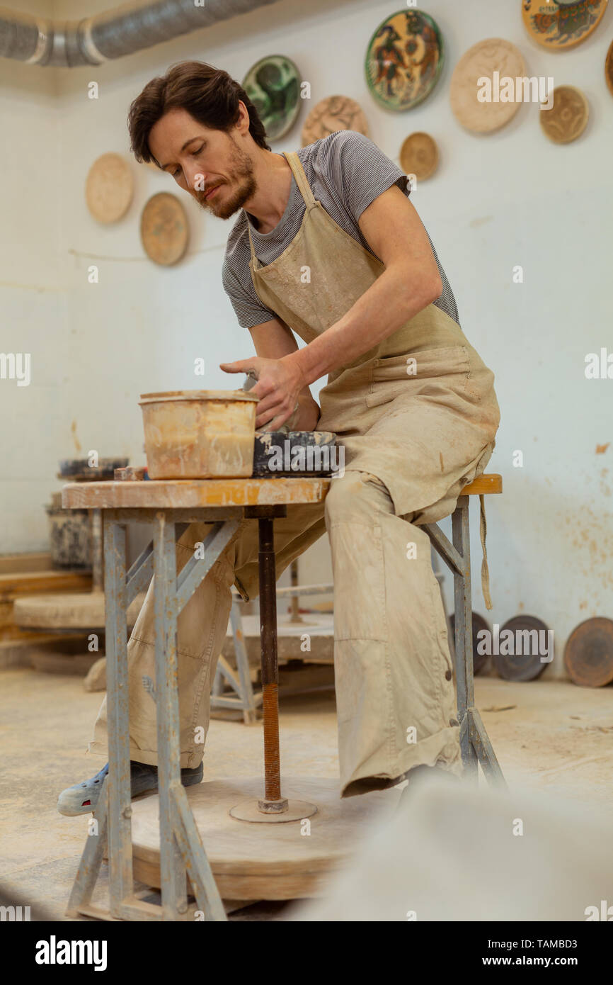 Dealing with clay. Bearded experienced man in grey apron sitting on ...