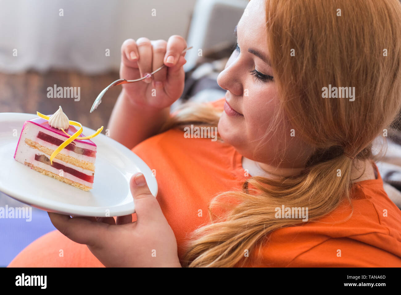 Chubby woman sport at home sitting eating cake concentrated smiling ...