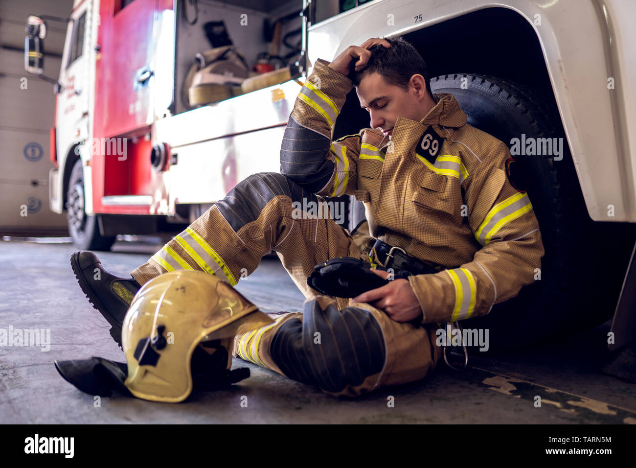 Photo of tired fireman sitting on floor near fire truck at station ...