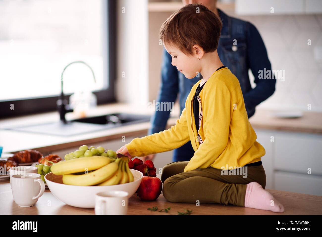 A small girl sitting on the counter in a kitchen, eating fruit Stock ...