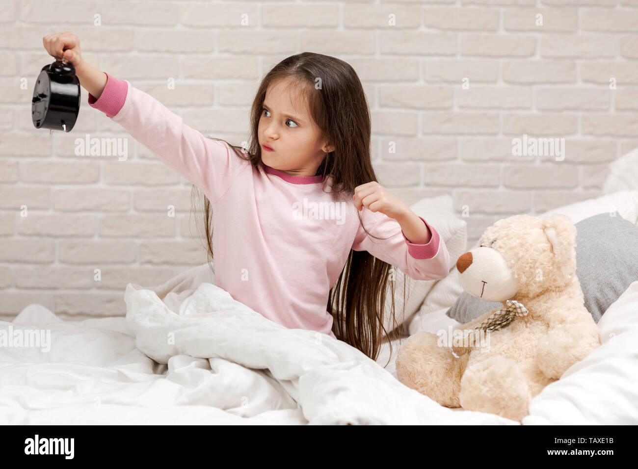 angry little child girl in pyjamas with clock in bed. good morning ...