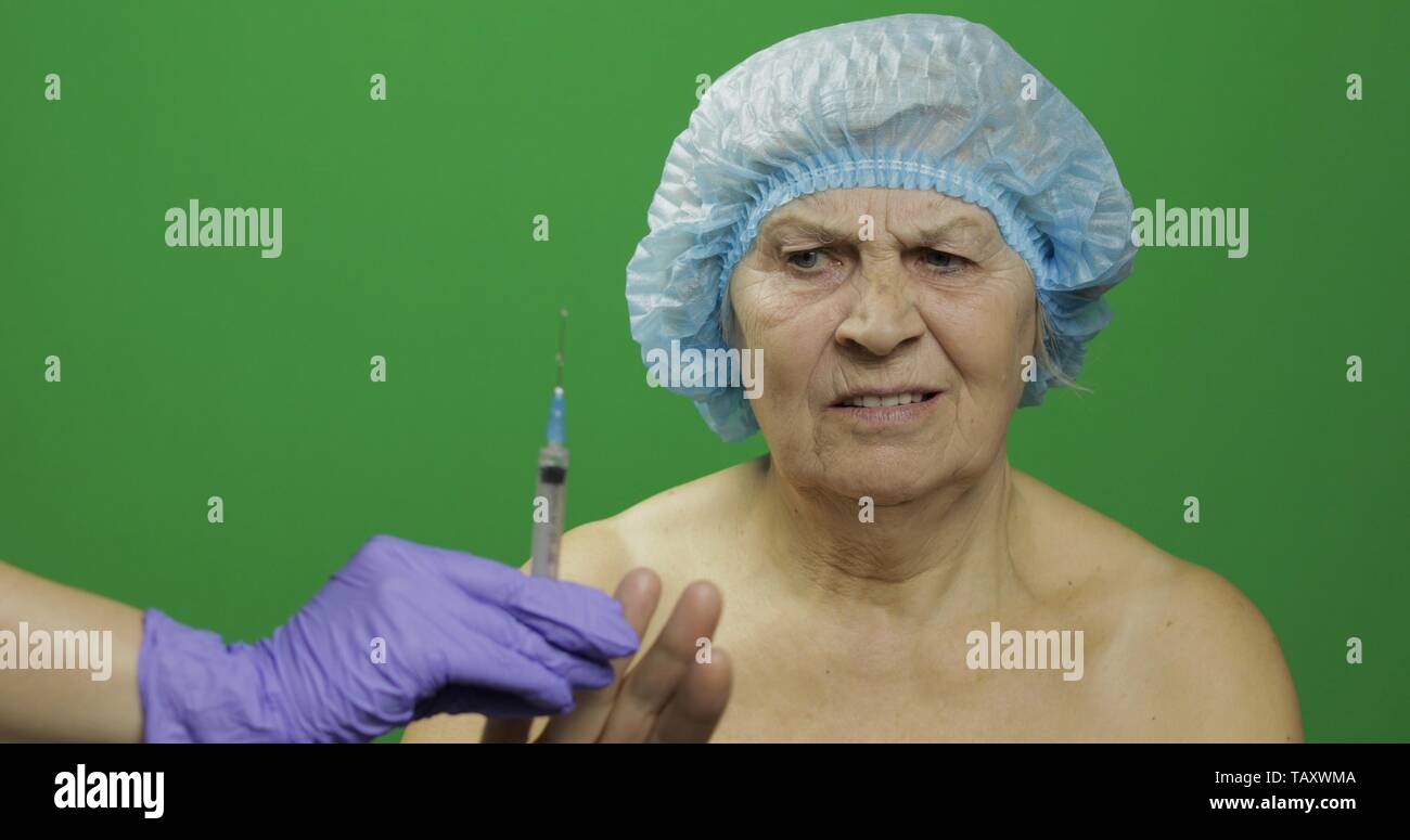 Smiling elderly female lady in protective hat scared of syringe with ...
