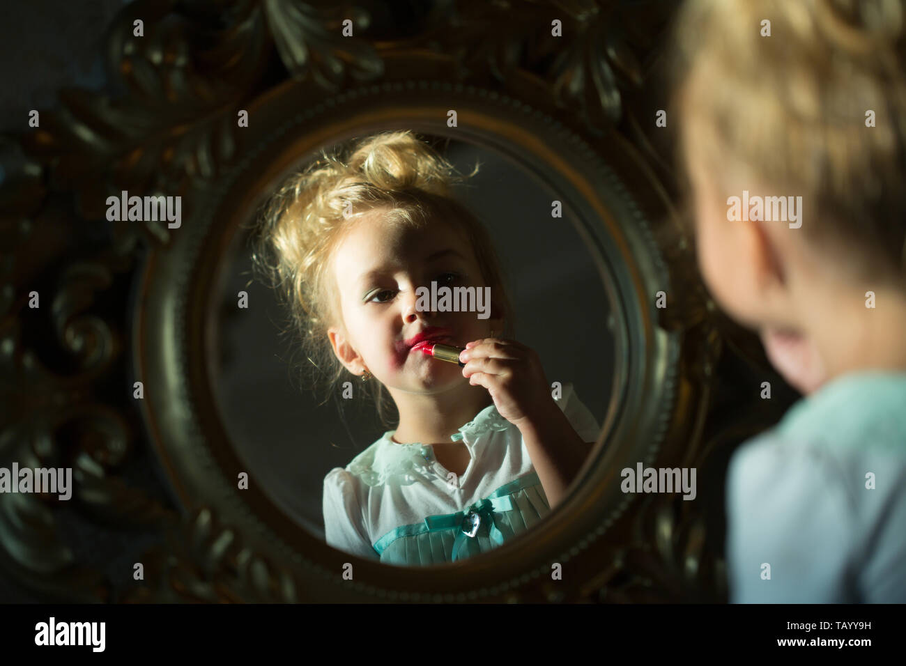 Baby girl put red lipstick on lips. Kid model make up at mirror in room ...