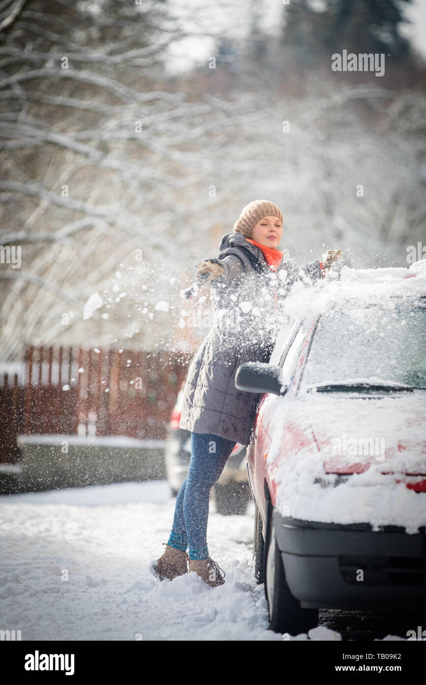 Pretty, young woman cleaning her car from snow after heavy snowstorm ...