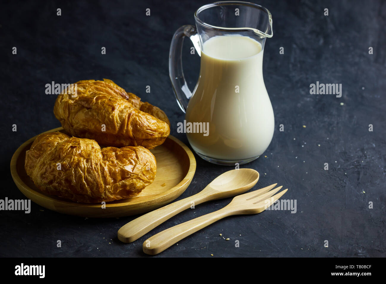 Croissants on wooden dish and milk jug on table in dark background ...