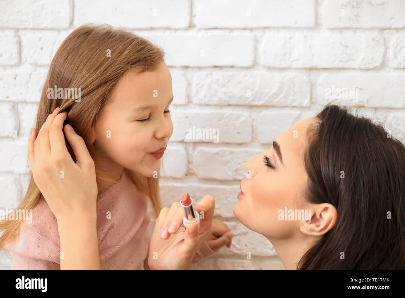 Young mother applying lipstick onto daughter's lips against white brick ...