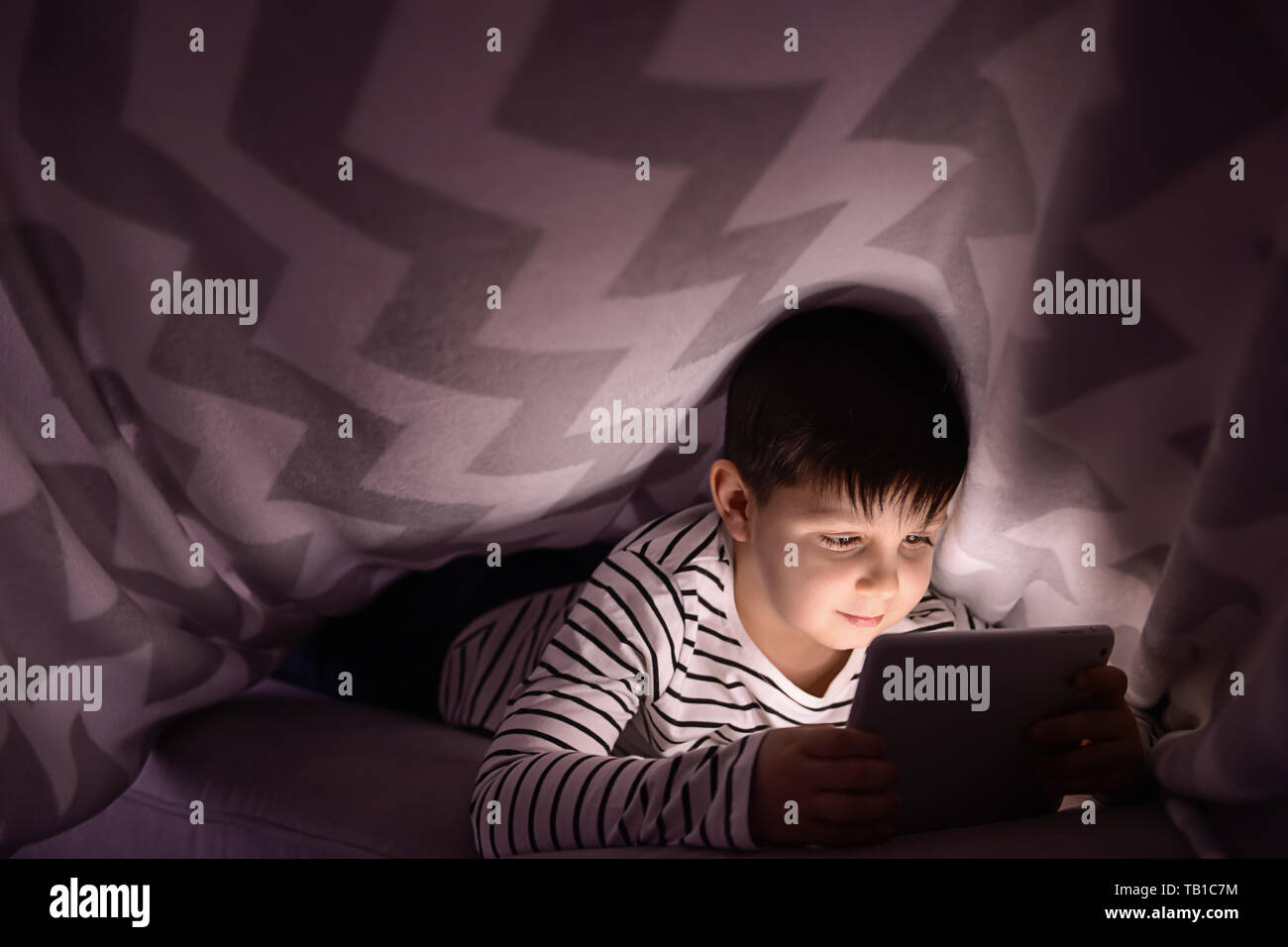 Cute little boy with tablet computer watching cartoons under blanket ...