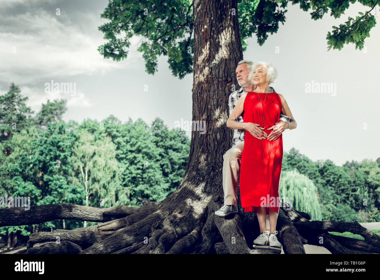 Attractive man tenderly cuddling wife while standing under the tree ...