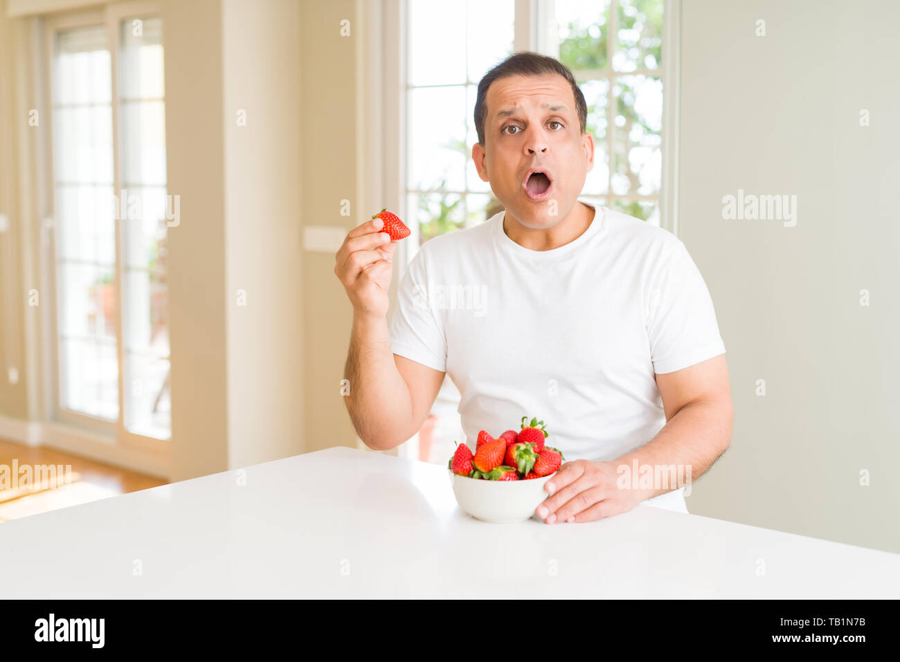 Middle age man eating strawberries at home scared in shock with a ...