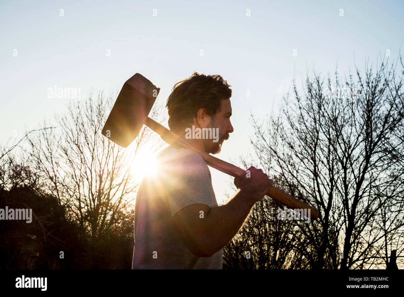 Bearded man walking outdoors at sunset, carrying large wooden mallet on ...