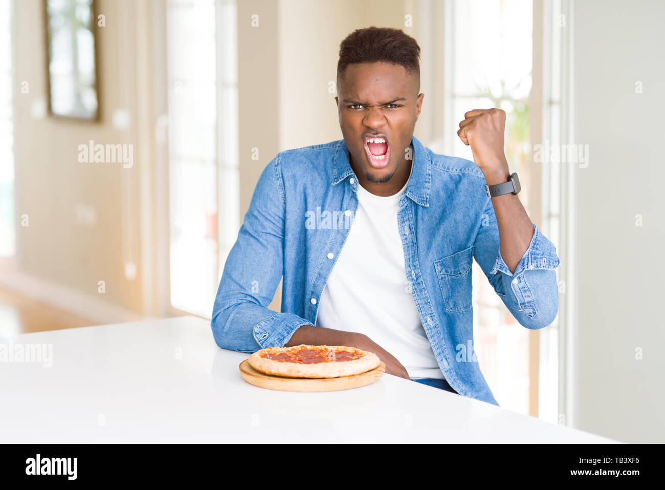 African american man eating pepperoni pizza at home annoyed and ...