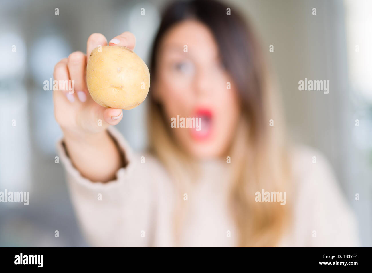 Young beautiful woman holding fresh potato at home scared in shock with ...