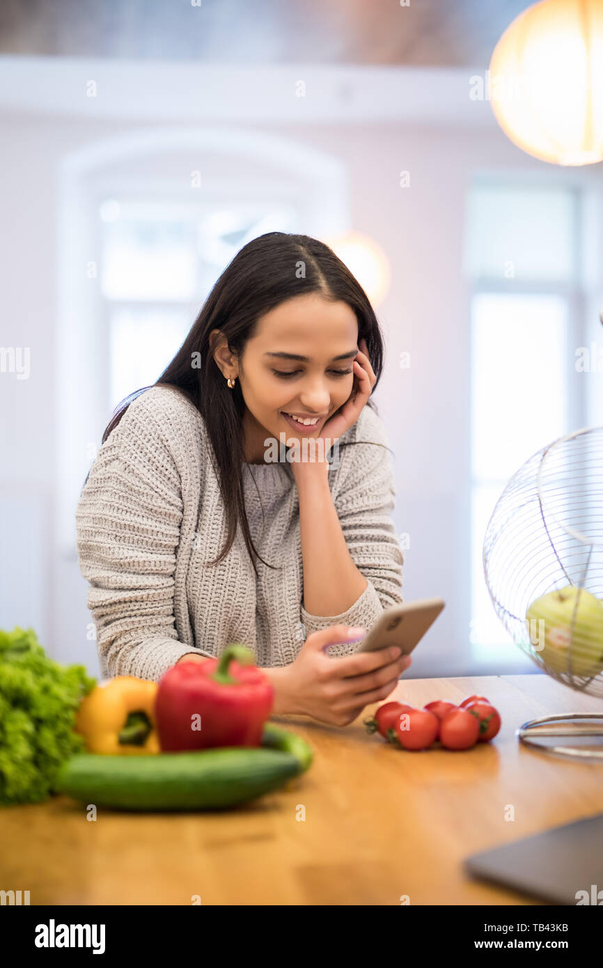 Smiling woman text messaging in front of vegetables in the kitchen at ...