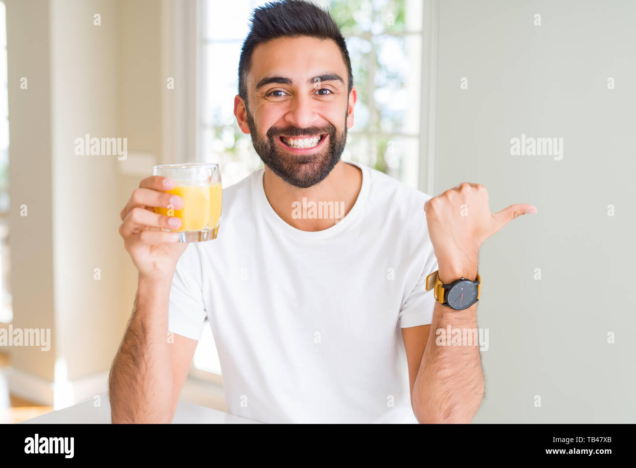 Handsome hispanic man drinking healthy orange juice pointing and ...