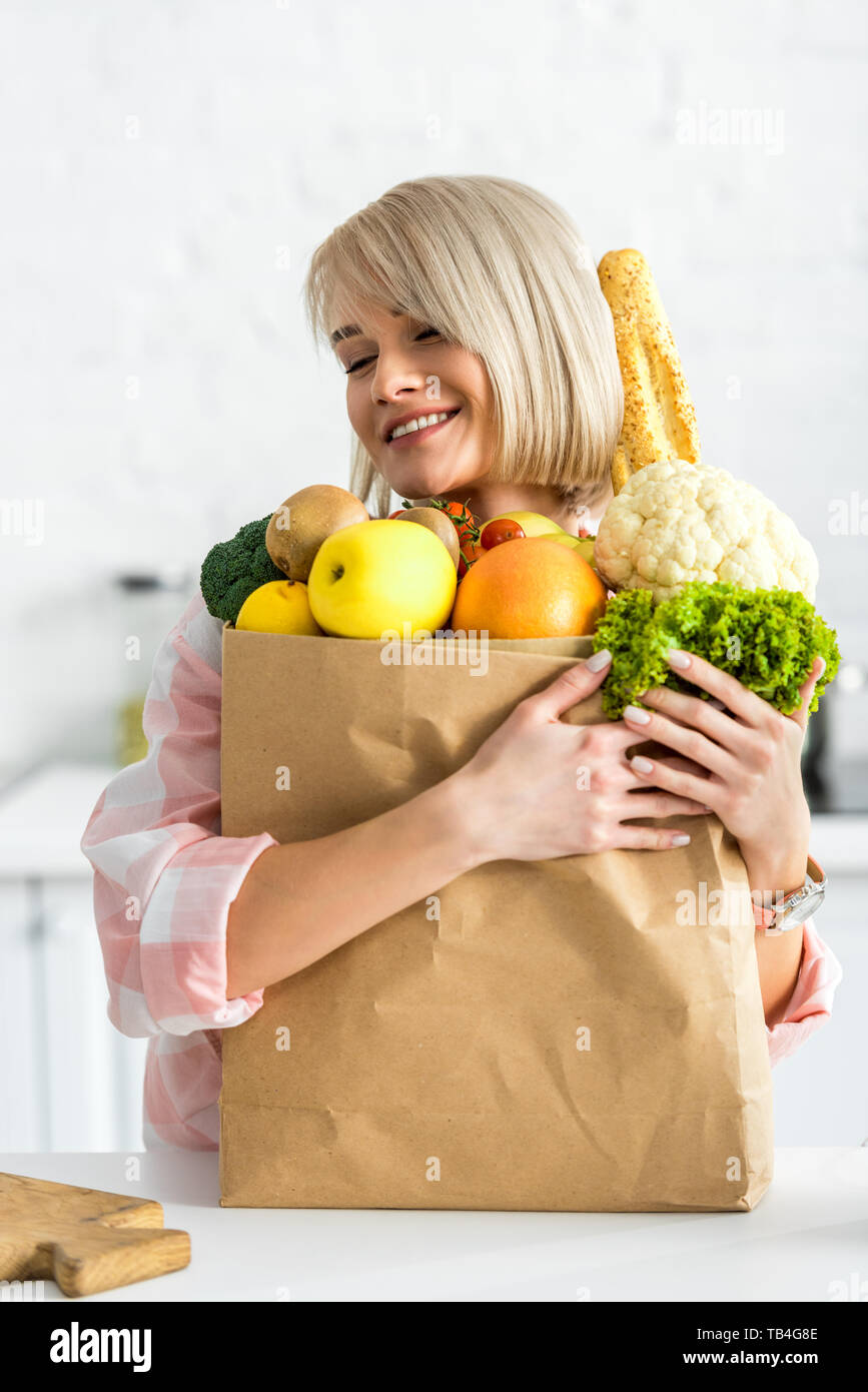 happy blonde young woman hugging paper bag with groceries Stock Photo ...