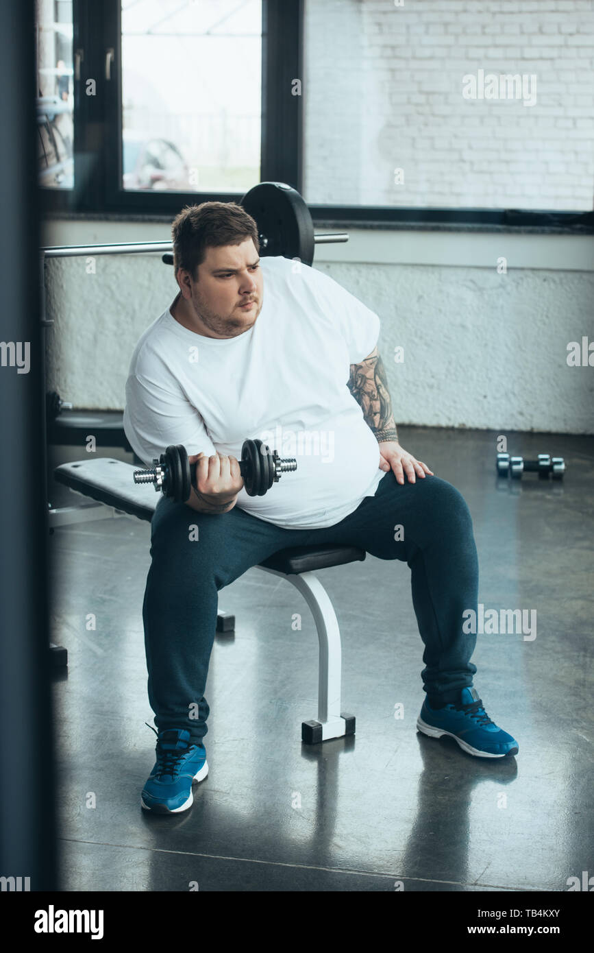 Overweight tattooed man sitting and exercising with dumbbell at sports ...