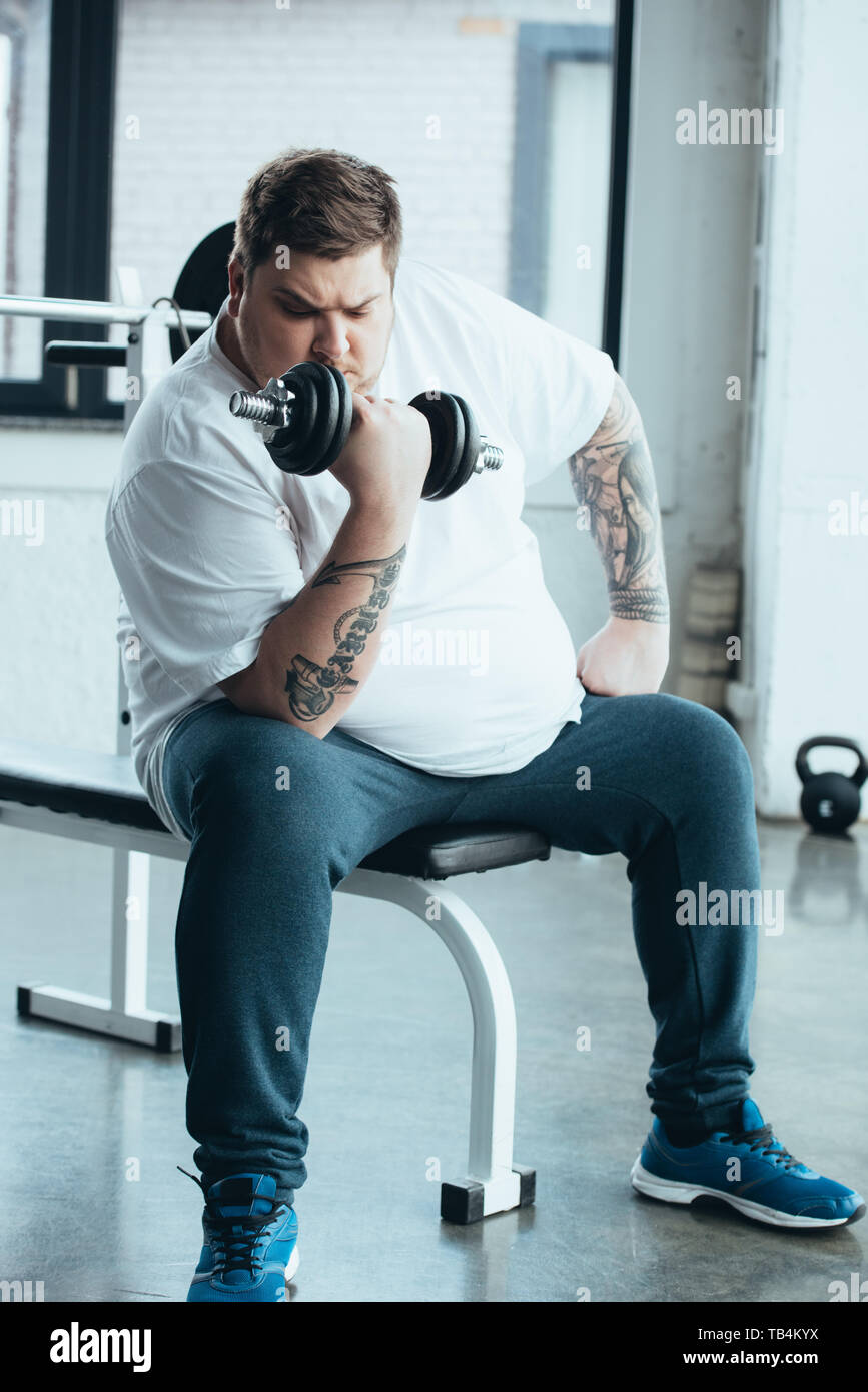 Overweight tattooed man sitting and training with dumbbell at sports ...