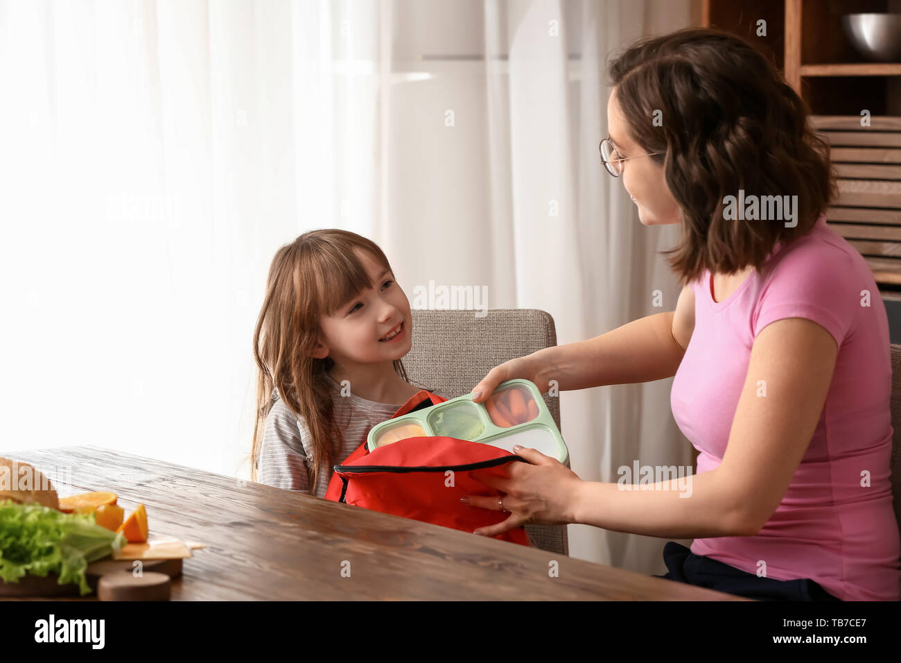 Mother with cute daughter packing lunch box into backpack at home Stock ...