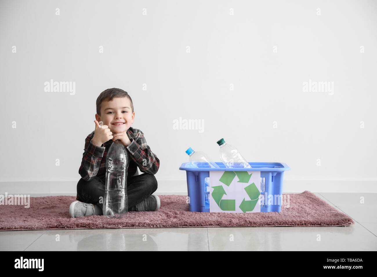 Little boy sitting on floor near basket for trash against white wall ...