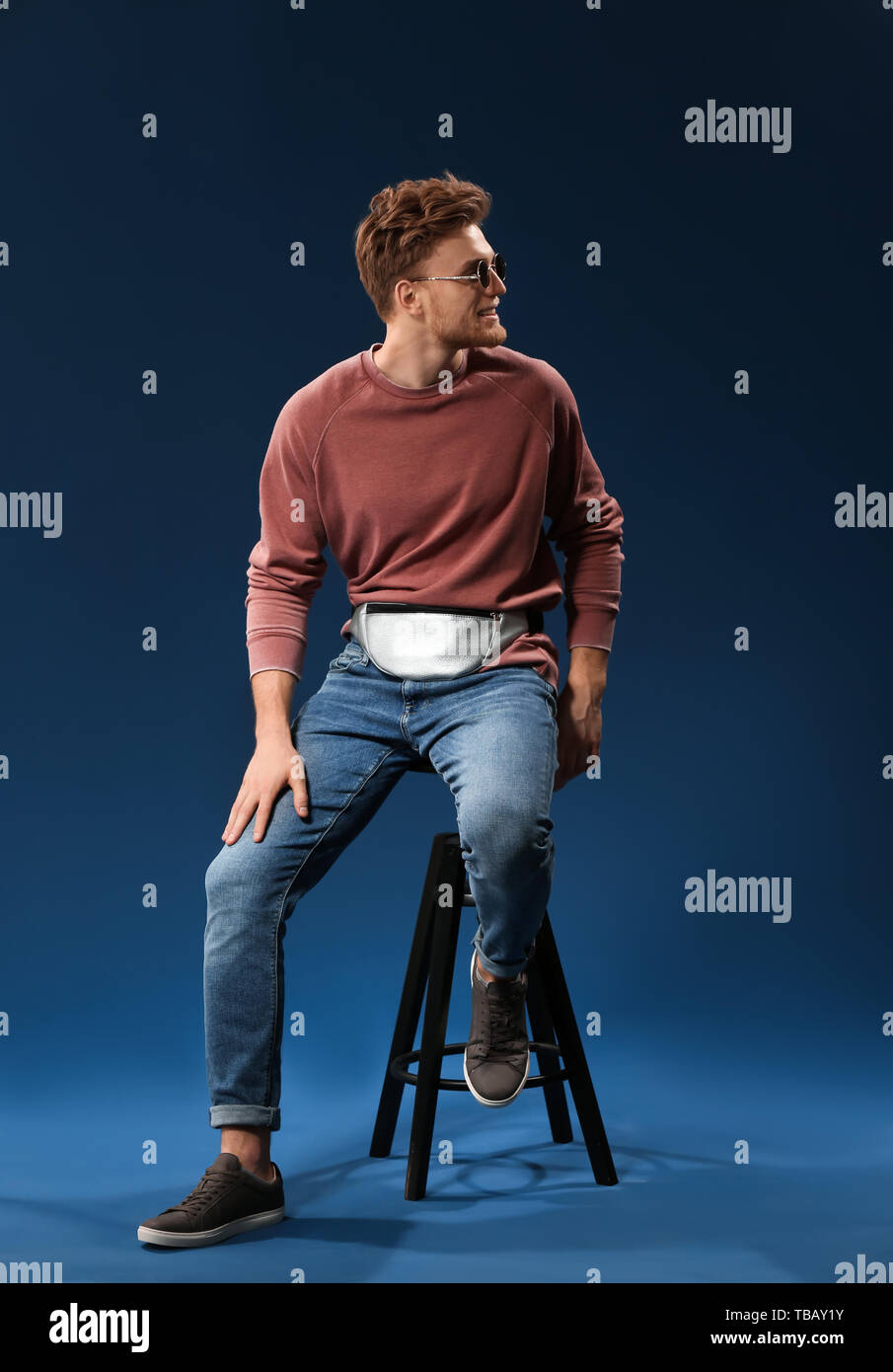 Fashionable young man sitting on stool against color background Stock ...