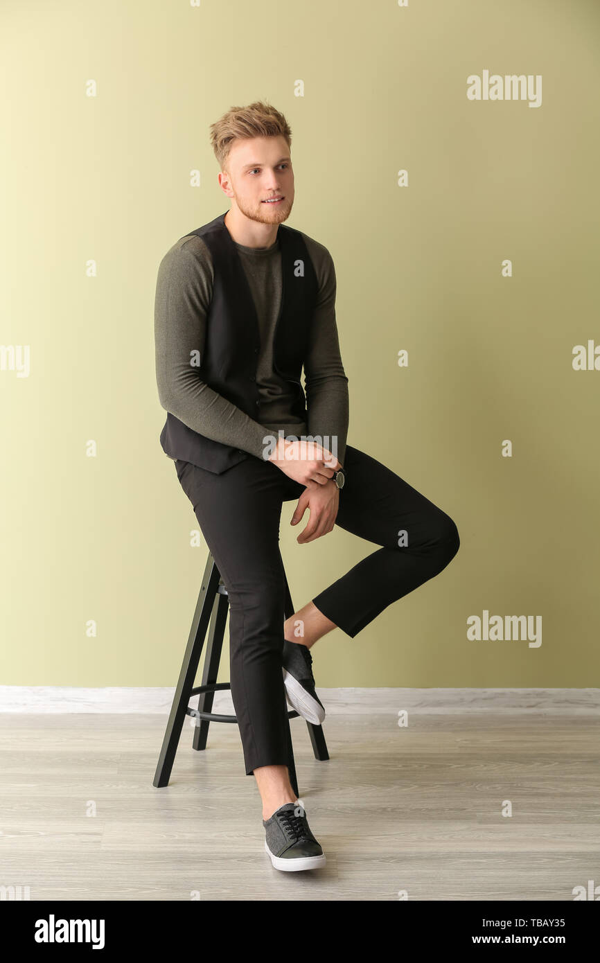 Fashionable young man sitting on stool against color wall Stock Photo ...