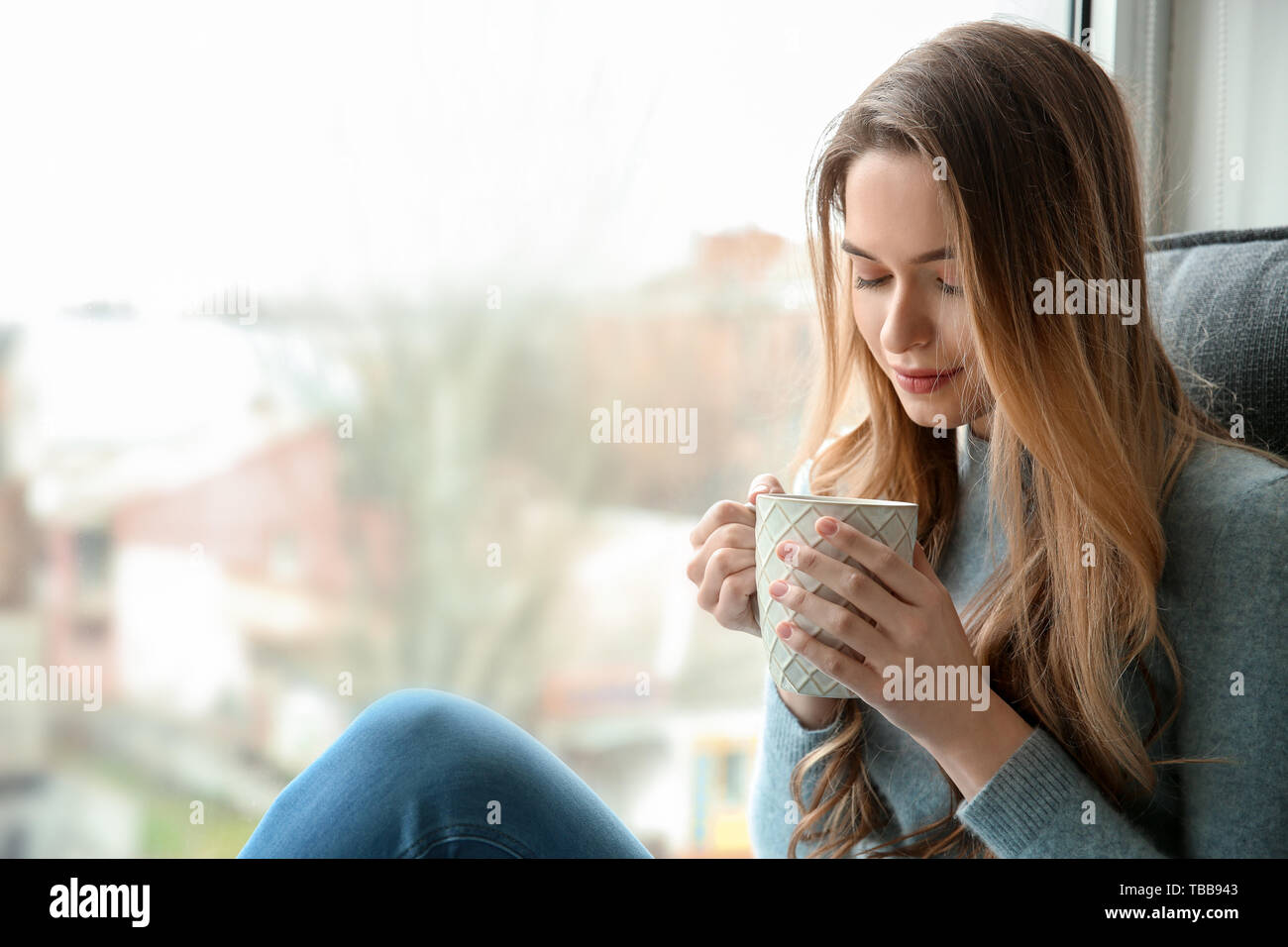 Beautiful young woman drinking coffee while sitting on window sill ...